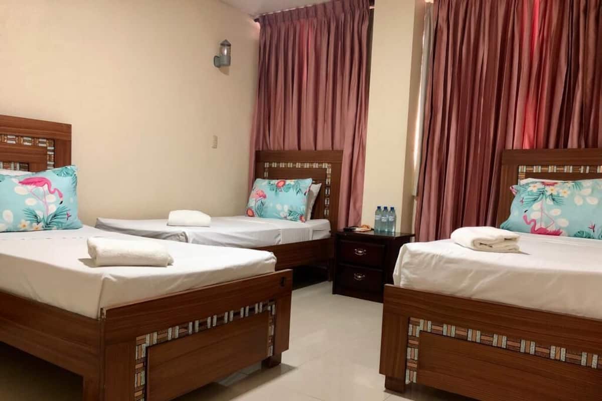 Quarto de hotel com duas camas de madeira, cada uma com almofada florida e manta branca. Há uma pequena mesa de cabeceira de madeira, com uma garrafa de água e um abajur.