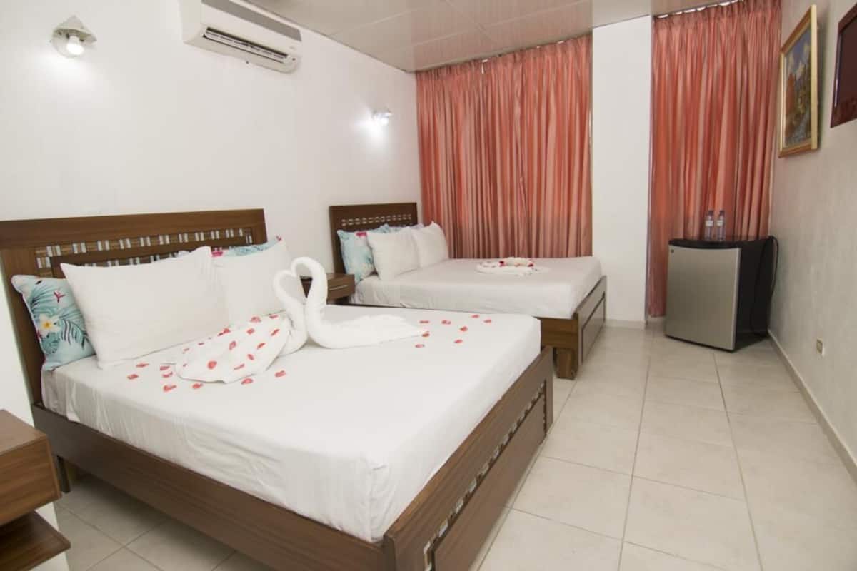 Quarto de hotel com duas camas, cada uma com cabeceira de madeira, roupa de cama branca e cisnes decorativos. Há uma pequena mesa com uma garrafa e um quadro na parede.