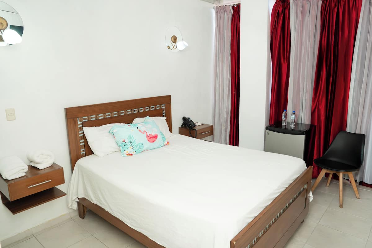 Cama bem arrumada com lençóis brancos, cabeceira de madeira e travesseiro floral. Um criado-mudo com abajur e telefone. O quarto possui uma pequena escrivaninha com cadeira e uma garrafa de água.