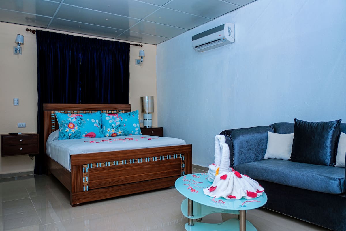 Quarto de hotel com cama de madeira, sofá azul, mesa redonda e unidade de ar condicionado na parede.