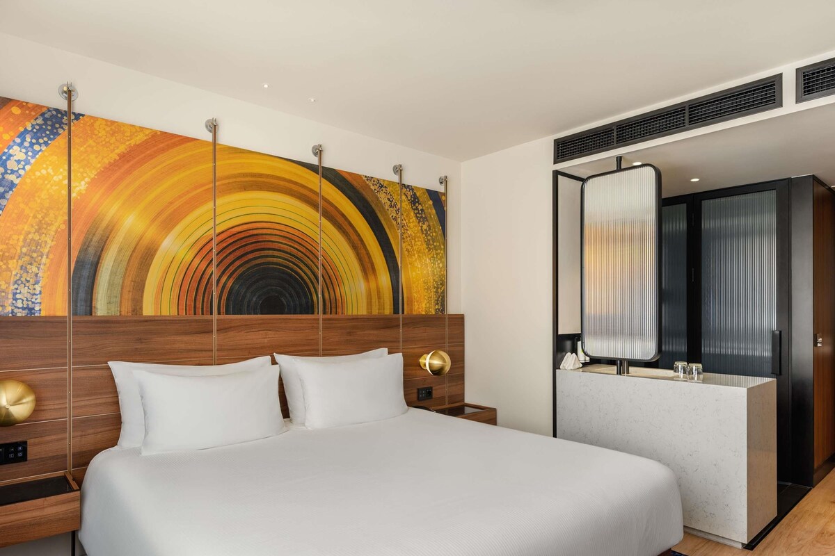 Quarto de hotel com uma cama grande, cabeceira de madeira, pintura abstrata colorida, banheiro moderno com espelho e uma pequena mesa de cabeceira.