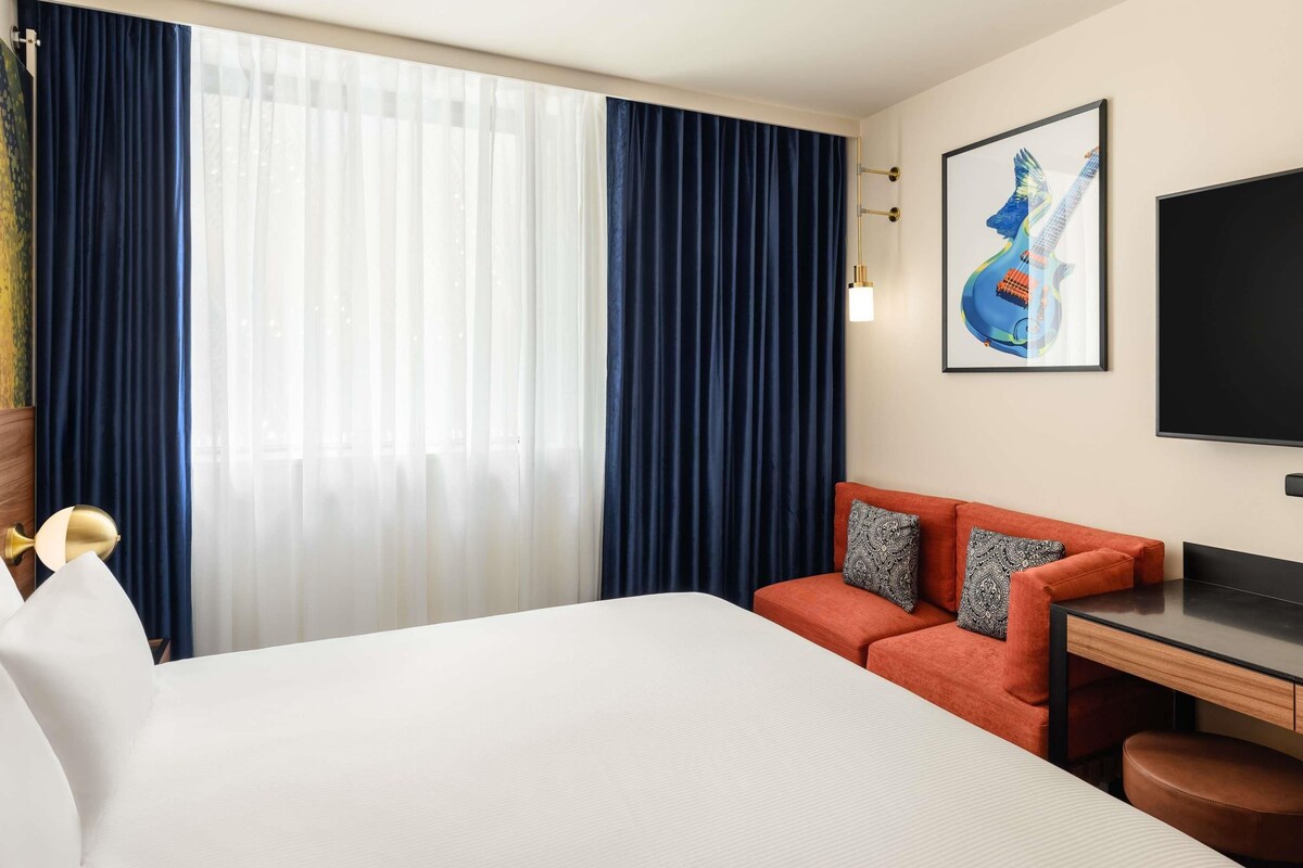 Quarto de hotel com cama, escrivaninha, cadeira, televisão e um quadro na parede.