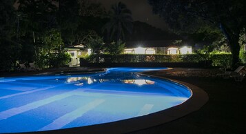 Piscina à noite com vegetação ao redor e um edifício ao fundo.