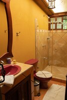 Casa de banho com parede amarela, toucador de madeira, lavatório vermelho, sanita e cabine de chuveiro em vidro.