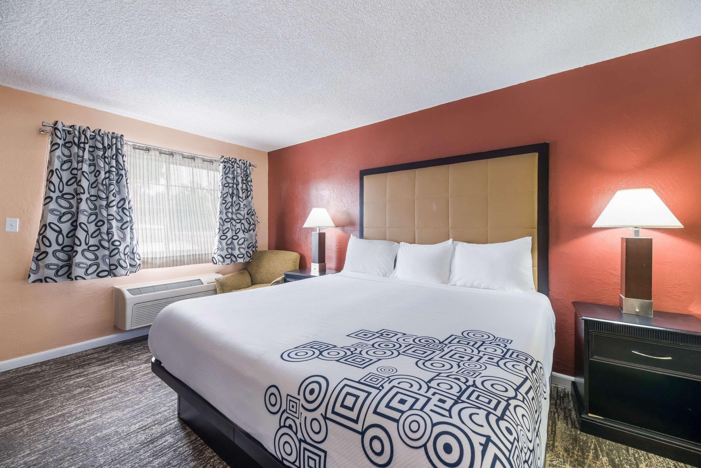Quarto de hotel com uma cama grande, uma luminária de cabeceira, uma cadeira e uma janela com cortinas estampadas.