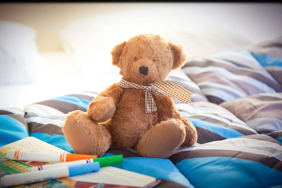 Um urso de pelúcia sentado em uma cama com roupa de cama listrada e diversos materiais de papelaria.