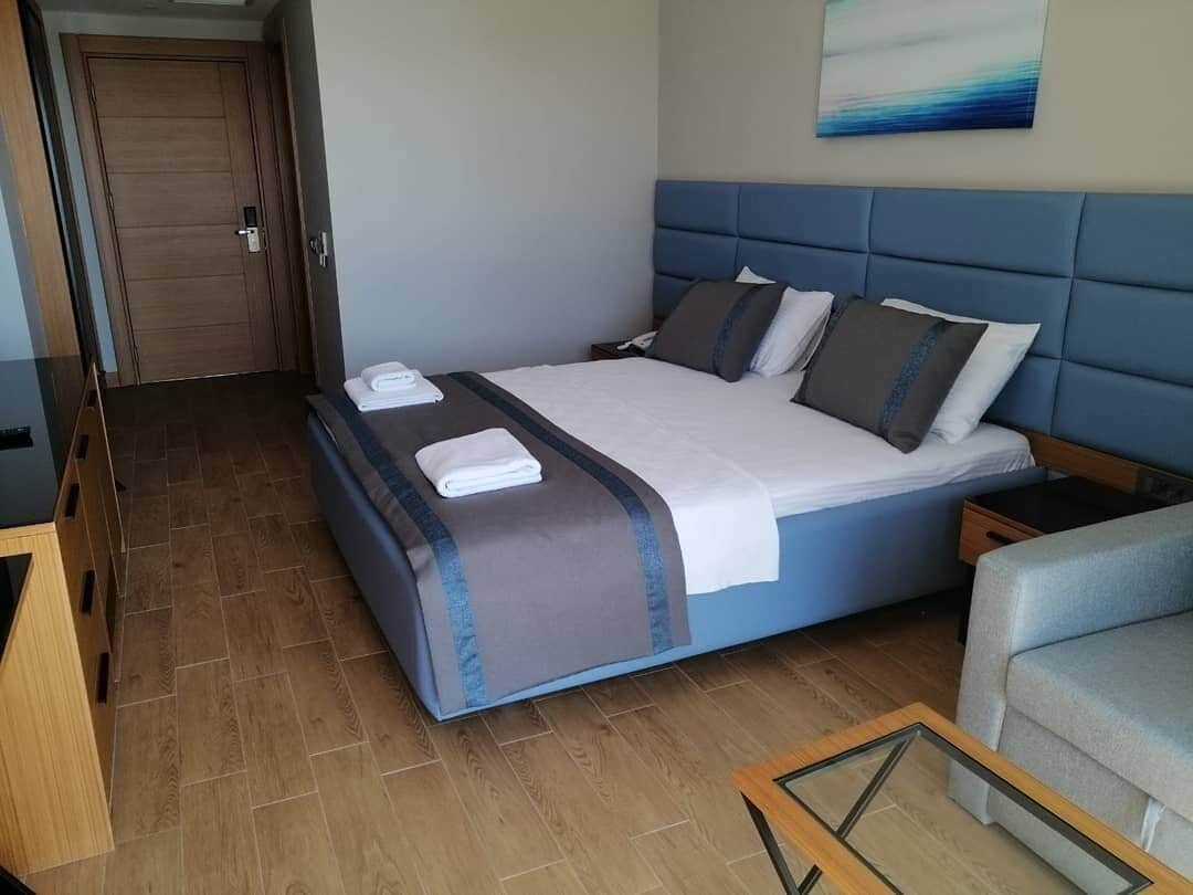 Uma cama bem arrumada com cabeceira azul, uma porta de madeira e uma pequena mesa com tampo de vidro.