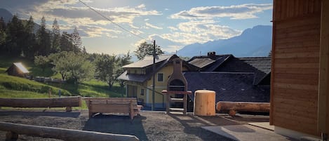 Cenário rural com um prédio de madeira, um banco e uma trilha de terra que leva a uma casa com telhado de telhas.