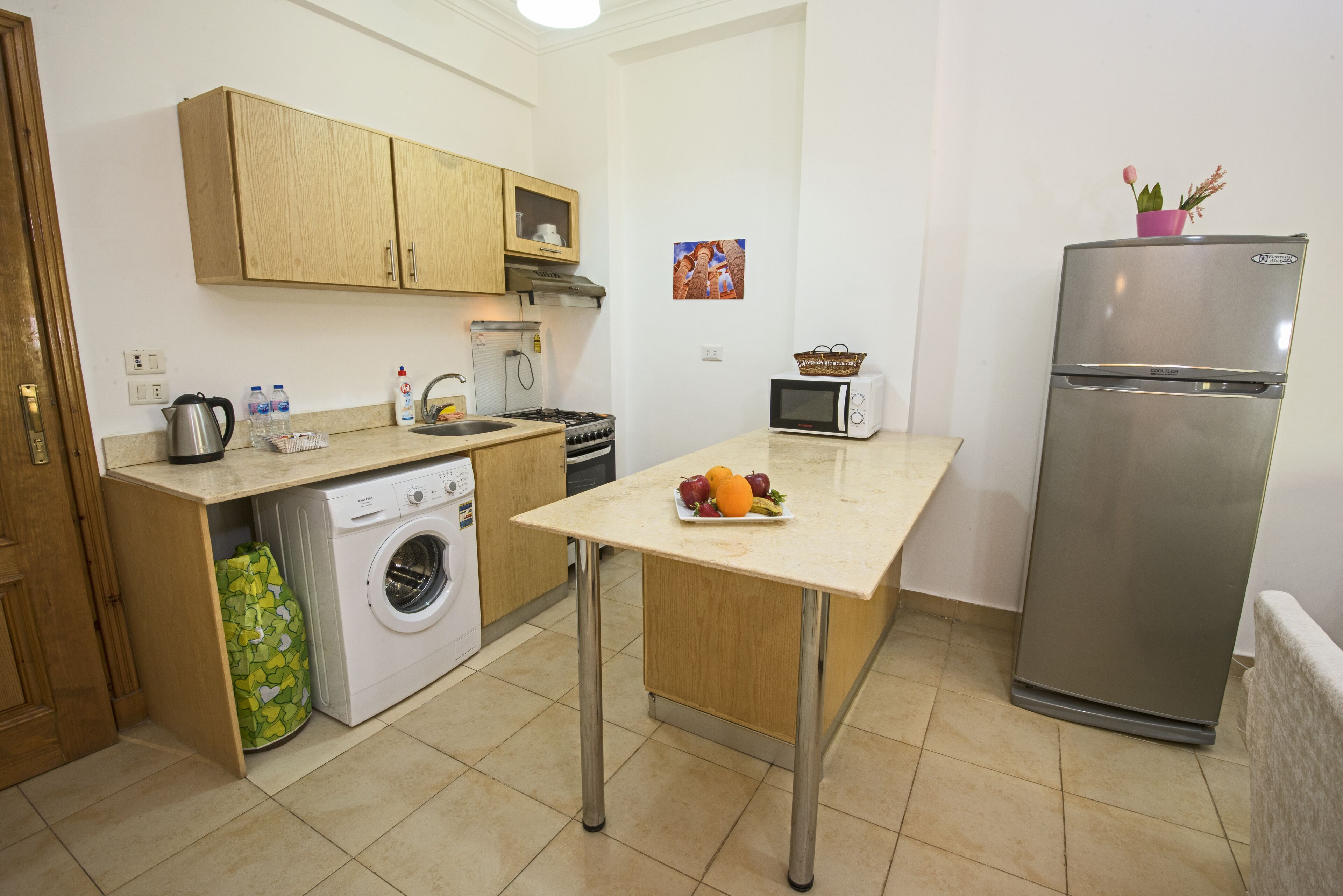 Cozinha compacta com armários de madeira, geladeira de aço inoxidável, micro-ondas e máquina de lavar.
