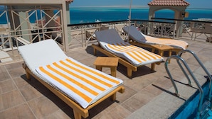 Área à beira da piscina com espreguiçadeiras e guarda-sóis, com vista para um mar azul e límpido.