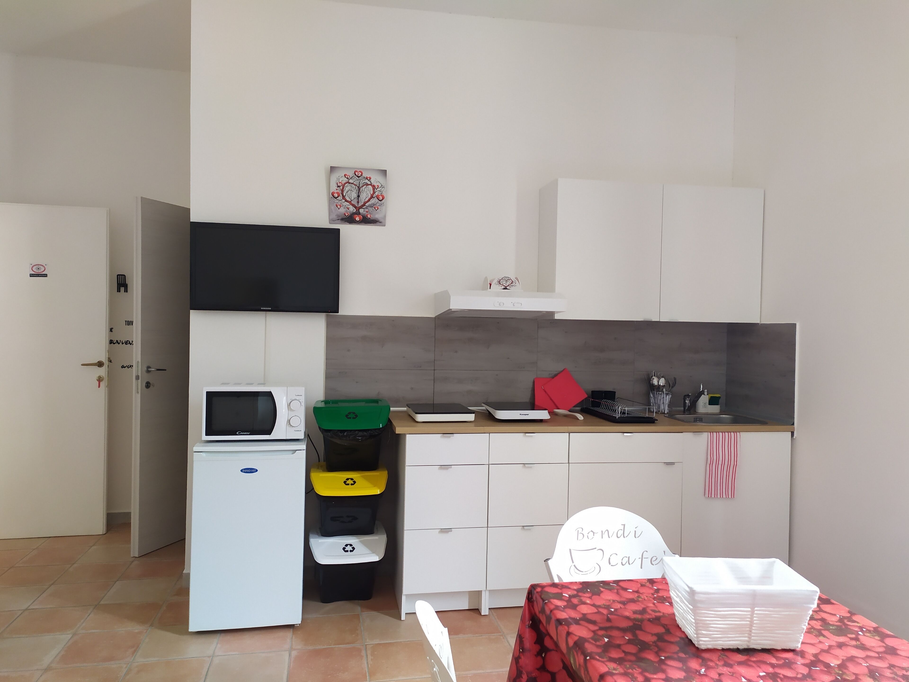 Cozinha compacta com micro-ondas, mini geladeira e pia. Há uma mesa com toalha vermelha e um letreiro que diz "Bondi Cafe".