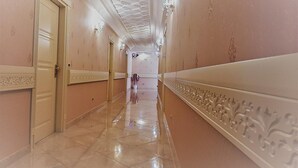 Um corredor com detalhes ornamentais nas paredes, piso ladrilhado e porta com a inscrição '102'.