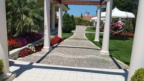 Um caminho pavimentado com colunas brancas, cercado por canteiros de flores e vegetação.