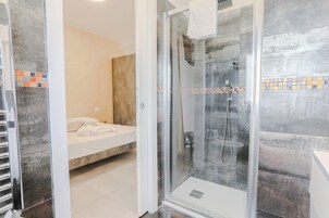Casa de banho moderna com cabine de chuveiro em vidro, sanita visível e cama no quarto adjacente.