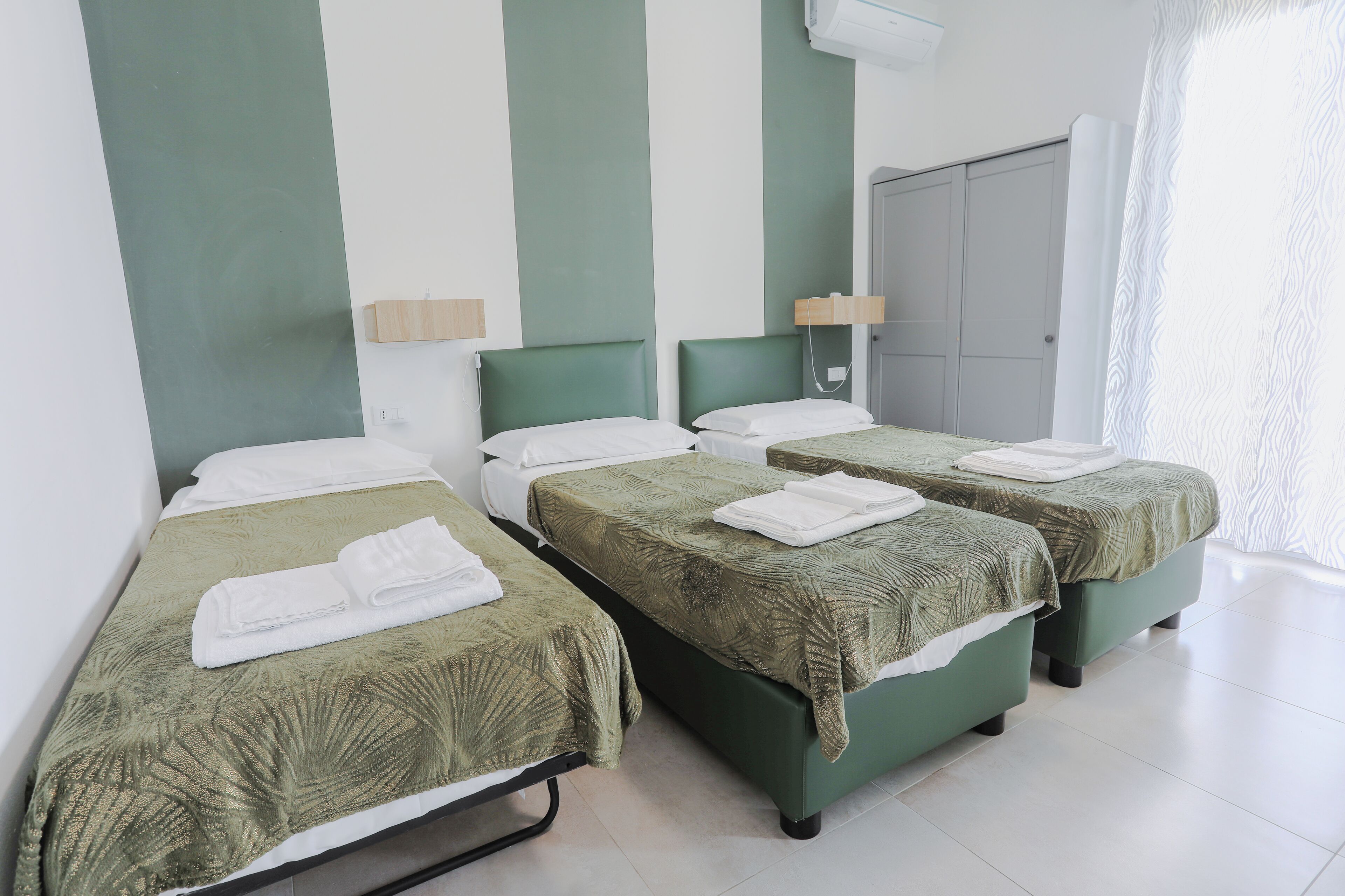 Quarto de hotel com três camas individuais, cada uma com cabeceira verde e roupa de cama branca. Há duas luminárias de mesa e um guarda-roupa ao fundo.
