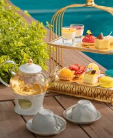 Uma mesa com um conjunto de chá, uma bandeja de sobremesas e vista para o mar.