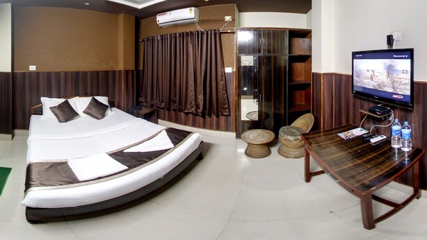 Quarto de hotel com cama curva, televisão, mesa pequena com controles remotos e garrafa de água.