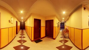 Corredor de hotel com padrão de piso xadrez, várias portas e um espaço de estar com um banco estampado.