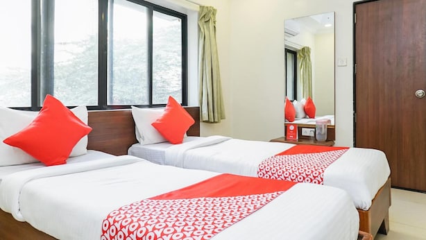 Quarto de hotel com duas camas, cada uma com roupa de cama branca e almofadas vermelhas. Há uma janela grande com cortinas, uma porta de madeira e um espelho na parede.