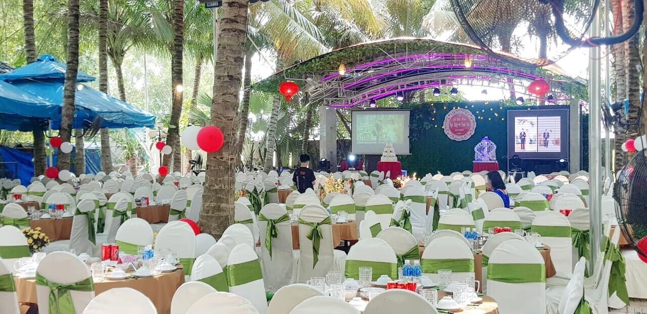 Evento ao ar livre com mesas e cadeiras dispostas em fileiras, um palco com tela e cenário tropical com palmeiras.