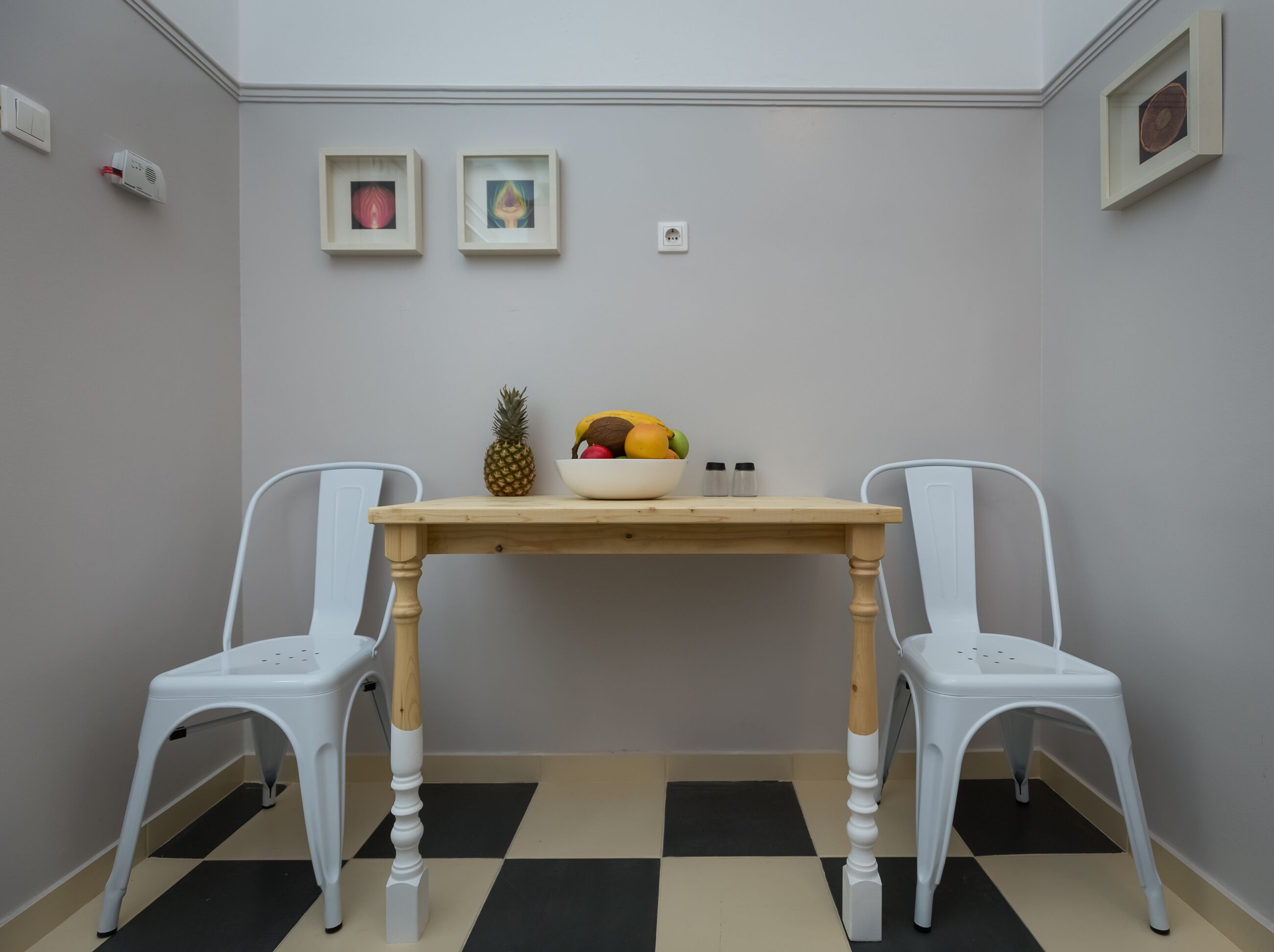 Quarto com piso xadrez, mesa de madeira, duas cadeiras brancas, uma fruteira e dois quadros na parede.
