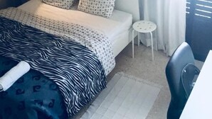 Quarto com cama, escrivaninha, cadeira, mesinha e janela com cortinas.