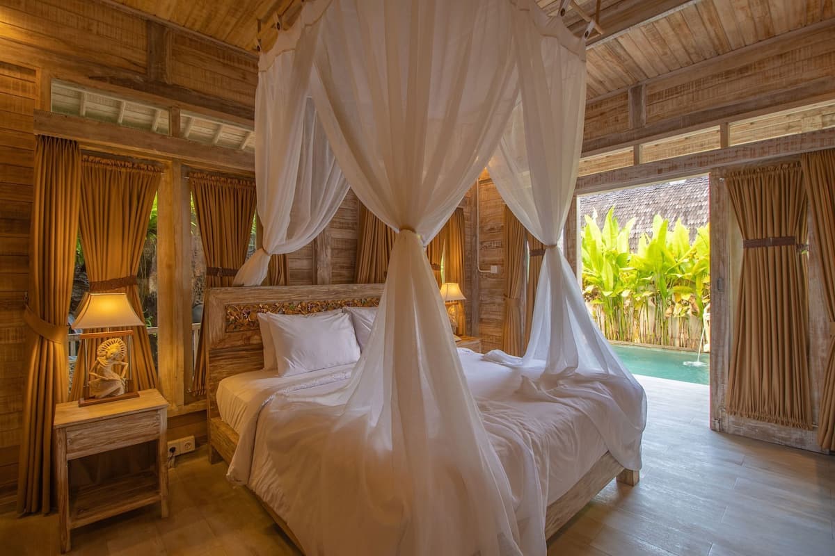 Uma cama de dossel com cortina, cabeceira de madeira, luminárias laterais e vista para uma piscina e plantas tropicais.
