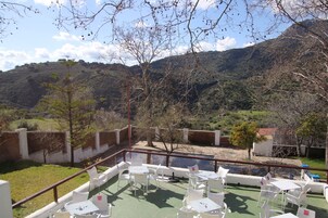 Terraço com mesas e cadeiras brancas, com vista para uma paisagem montanhosa e um corpo de água.