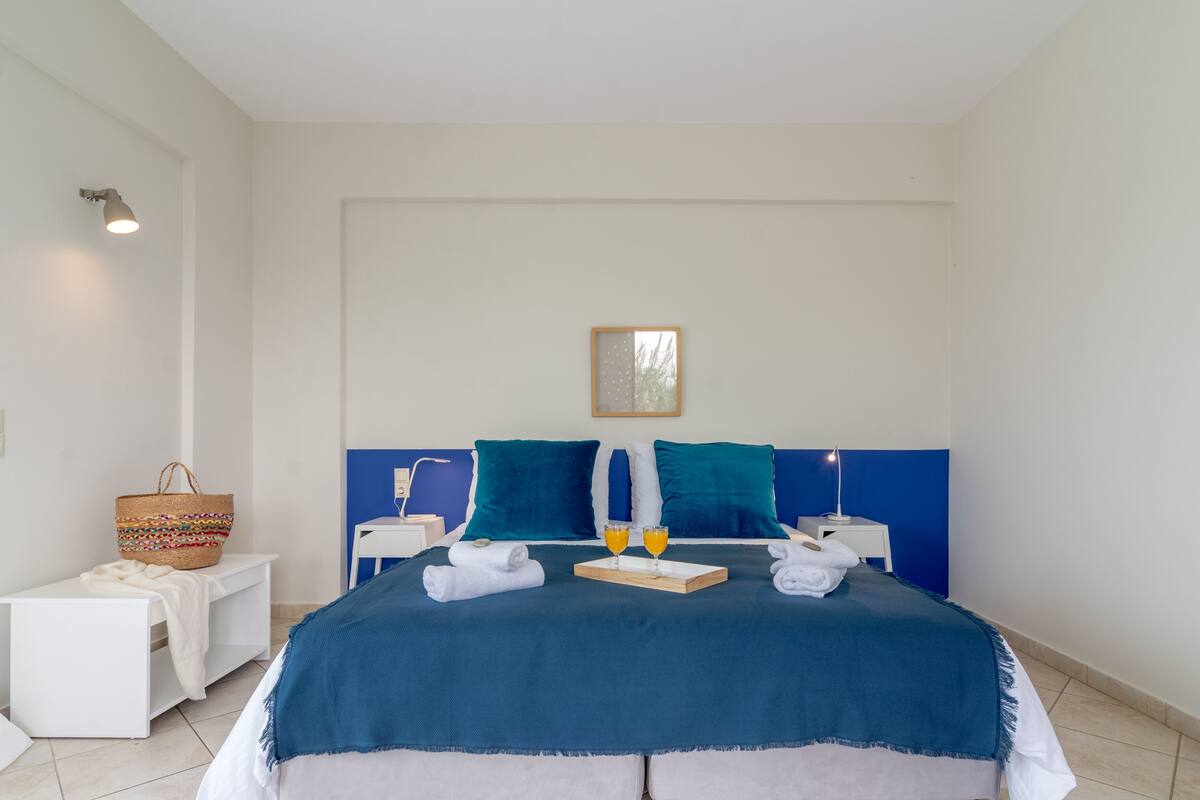 Cama bem arrumada com colcha azul, duas mesas de cabeceira com abajures, um cesto de tecido e toalhas.