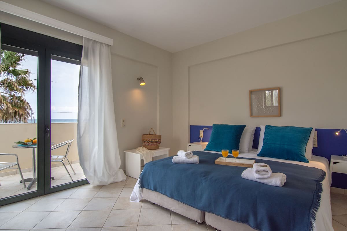Quarto de hotel com cama, uma mesinha com um cesto, uma cadeira e vista para o mar através de uma porta de correr.