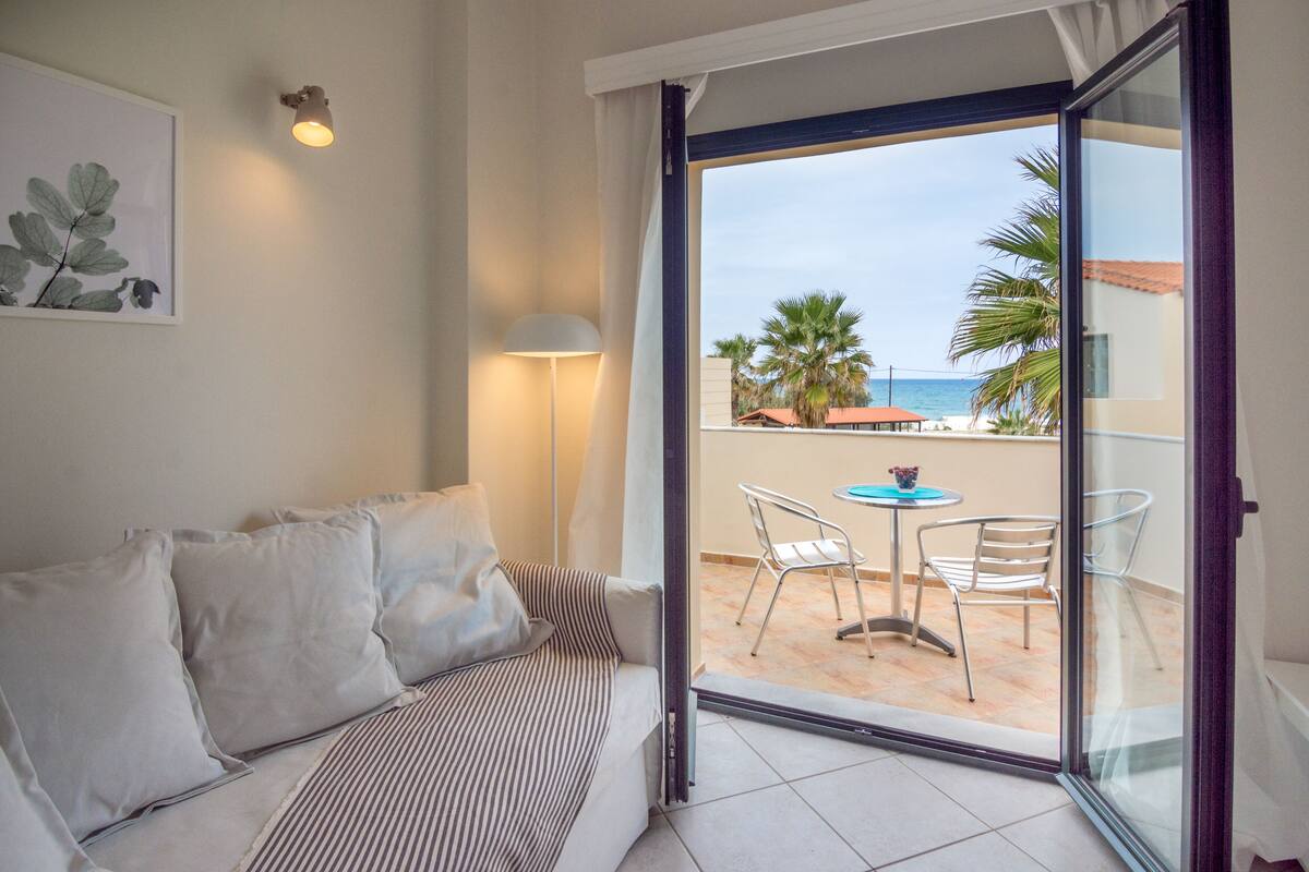 Uma varanda com sofá, mesa e cadeiras, com vista para uma praia com palmeiras e um prédio ao fundo.