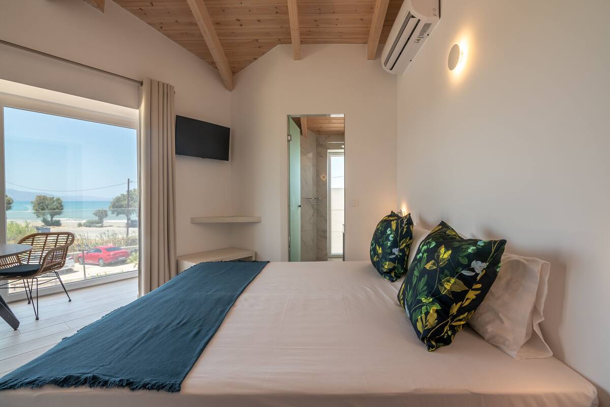 Quarto com cama, TV fixada na parede, janela com cortinas e vista para o exterior.