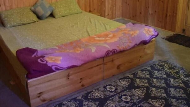 Uma cama de madeira com colcha verde e roxa, cabeceira de madeira e um tapete estampado no chão.