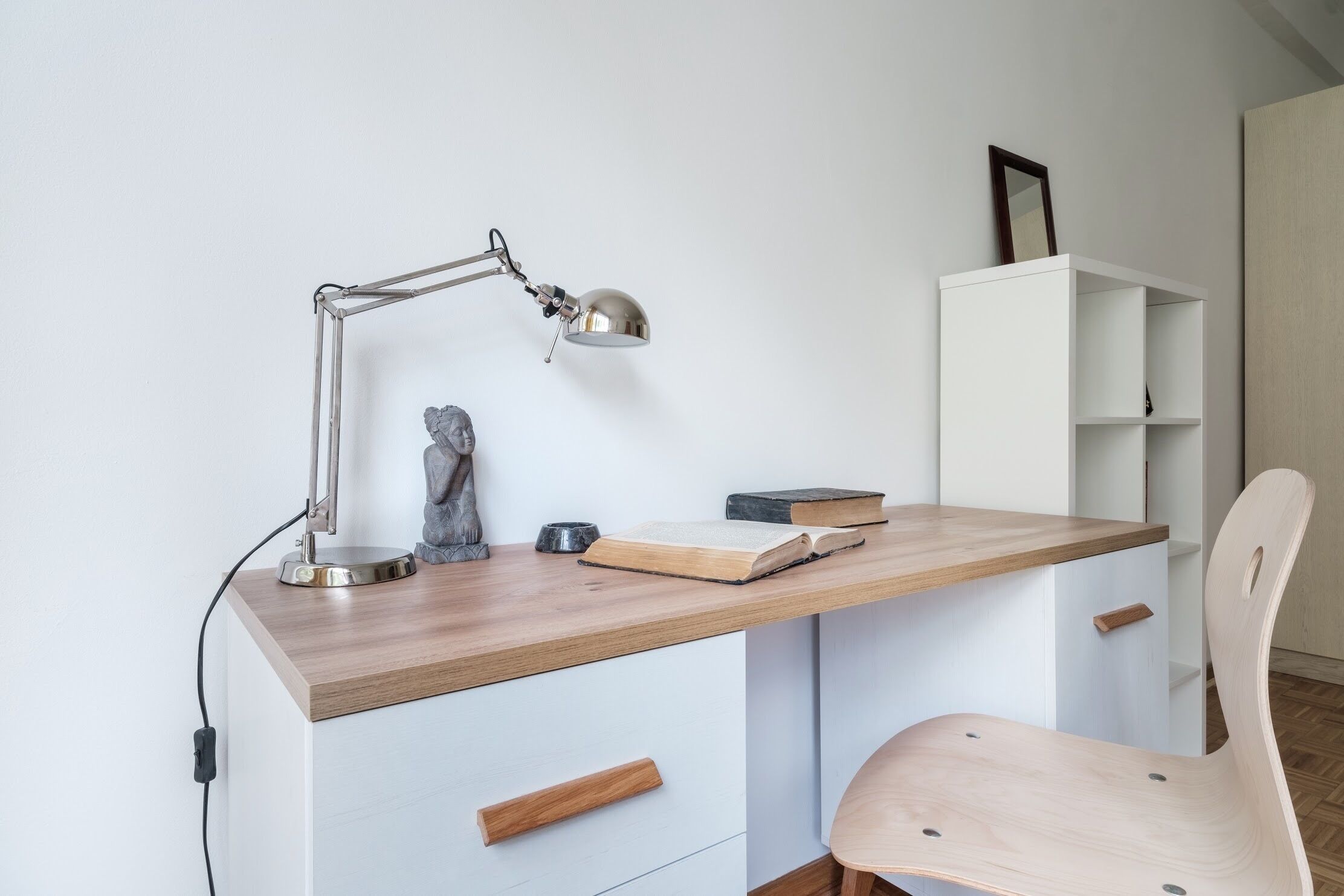 Uma mesa moderna com tampo de madeira, uma luminária de mesa metálica, uma cadeira e uma estante branca com um porta-retrato em cima.