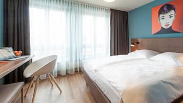 Quarto de hotel com cama, escrivaninha com laptop, cadeira e uma janela grande com cortinas.