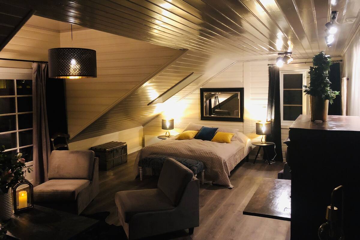 Quarto de sótão acolhedor com teto inclinado, uma cama grande, uma cadeira, uma mesa pequena e um vaso com planta.