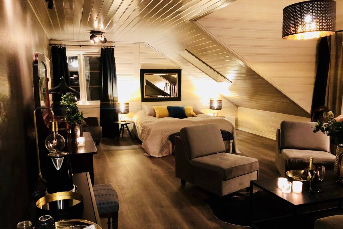 Um quarto aconchegante e bem iluminado, com uma cama, duas poltronas, uma mesinha e um espelho na parede.
