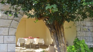 Uma área de refeições ao ar livre com uma parede de pedra, uma mesa posta com toalhas brancas e cadeiras, e uma árvore oferecendo sombra.