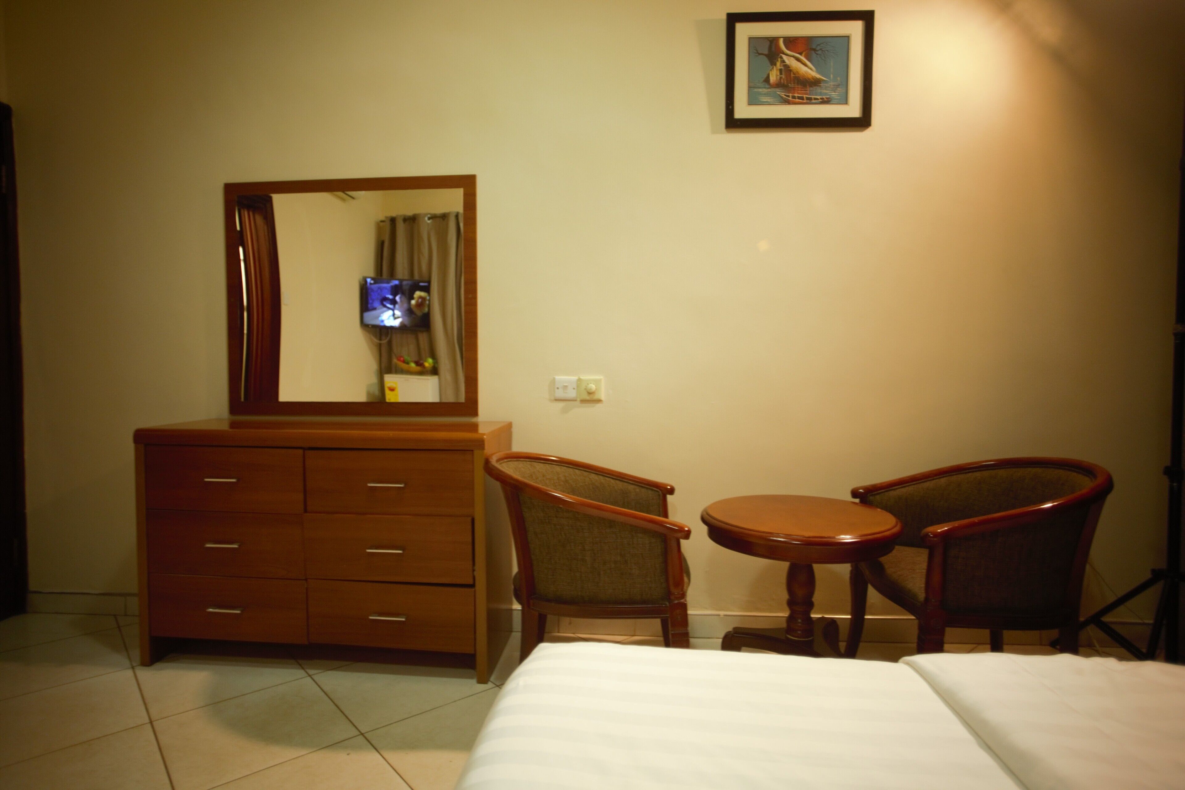 Quarto de hotel com cômoda de madeira, duas cadeiras, uma mesinha redonda, uma cama, televisão e um quadro na parede.