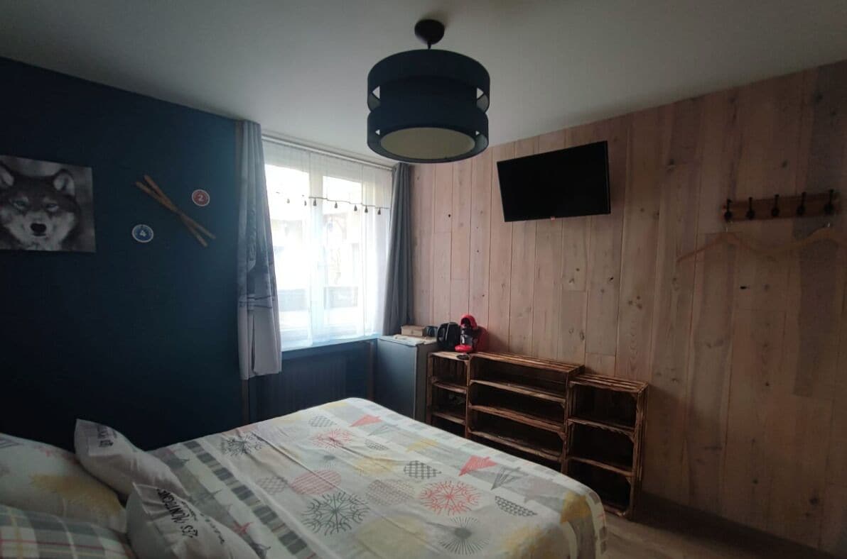 Quarto com cama, televisão na parede, cómoda de madeira e janela com cortinas.