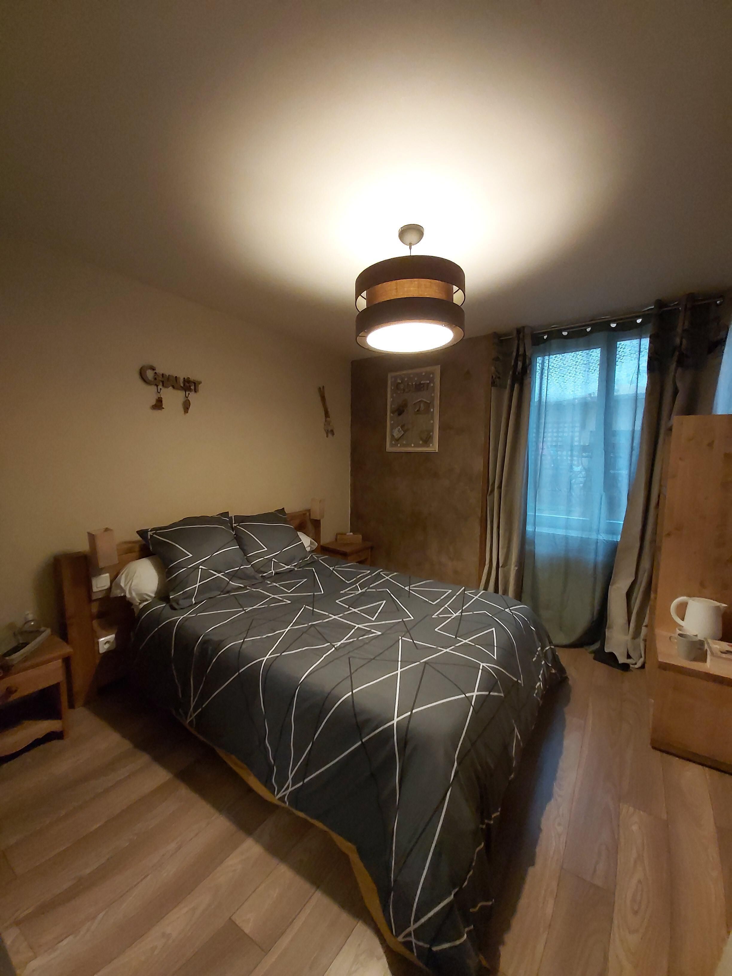 Quarto com cama, cabeceira de madeira, mesas de cabeceira, um criado-mudo, prateleira na parede, luminária pendente, janela com cortinas e um objeto decorativo na parede.