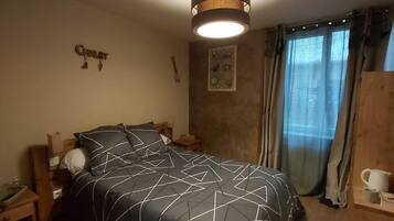 Quarto com cama, cabeceira de madeira, mesas de cabeceira, um criado-mudo, prateleira na parede, luminária pendente, janela com cortinas e um objeto decorativo na parede.
