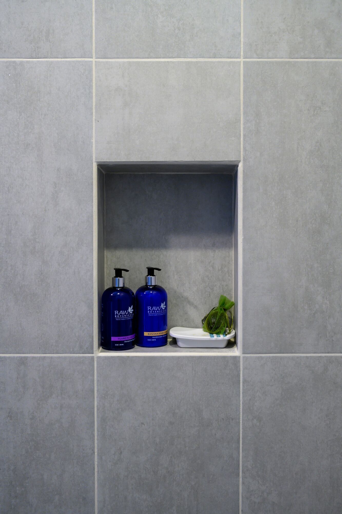 Um banheiro com azulejos cinza, uma prateleira com duas garrafas azuis identificadas como 'RAW BOTANICALS', e um pequeno recipiente branco com uma folha verde.