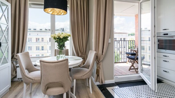 Uma área de jantar moderna com uma mesa redonda e quatro cadeiras, uma luminária pendente e vista da cidade através de grandes janelas.