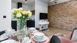 Uma área de jantar moderna com uma mesa redonda posta para quatro pessoas, um vaso de flores brancas e uma cozinha ao fundo.