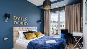 Quarto com cabeceira estofada, cama grande com roupa de cama azul e dourada, mesa de cabeceira e vista para edifícios pela janela.