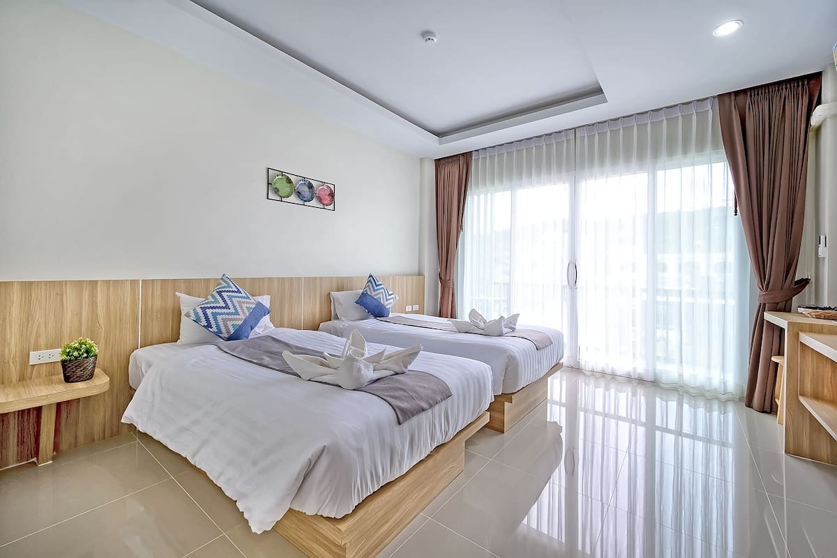 Quarto de hotel com duas camas, um banco, uma planta pequena e uma janela grande com cortinas.