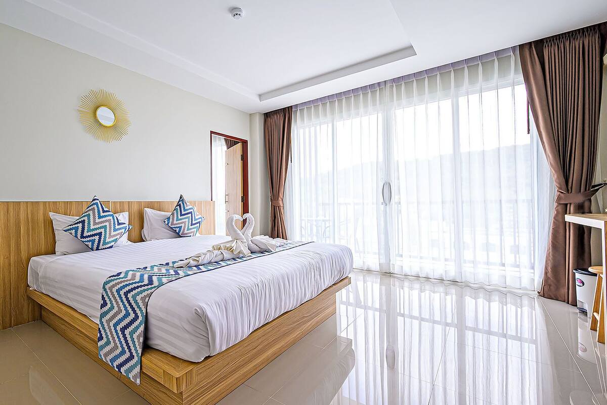 Quarto de hotel com cama grande, cabeceira de madeira e vista para o exterior através de cortinas leves.