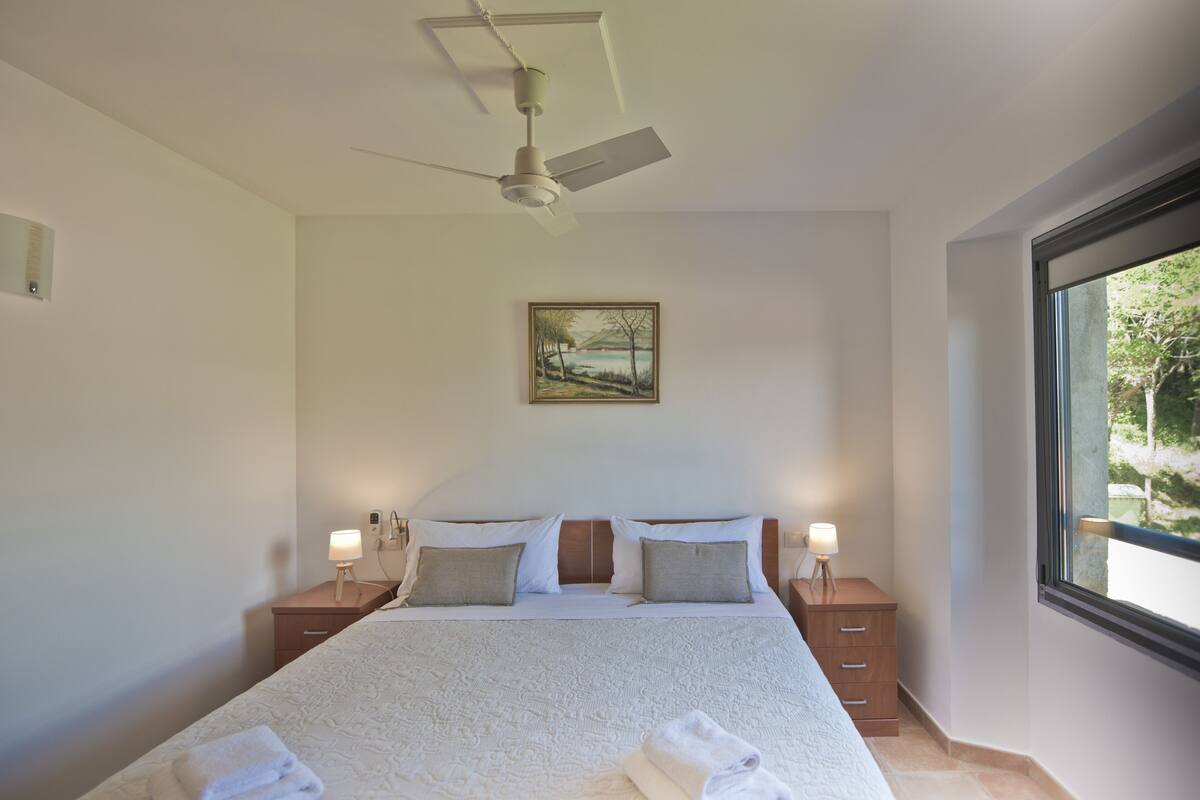 Um quarto com uma cama, duas mesas de cabeceira, um ventilador de teto, uma janela e um quadro na parede.