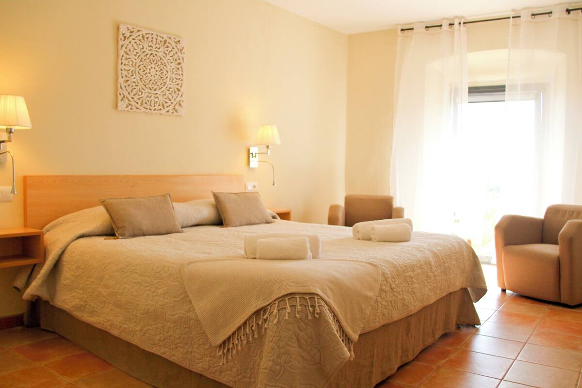 Quarto de hotel com uma cama grande, duas mesas de cabeceira, uma cadeira e uma janela com cortinas.